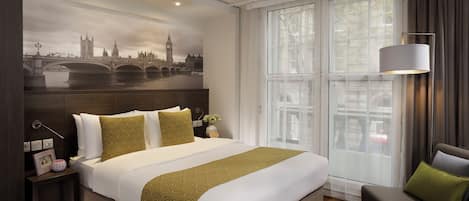 Quarto de hotel com uma cama grande, mesa de cabeceira, abajur, cadeira e uma janela ampla com vista para a paisagem urbana.