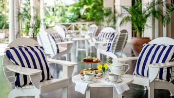 Mesa posta com xícaras de chá, um suporte de sobremesas e uma chaleira, cercada por cadeiras listradas em um ambiente de jardim interno.