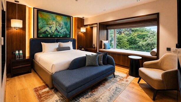 Quarto de hotel moderno com uma cama grande, um sofá, uma mesinha e uma janela com vista para o verde.