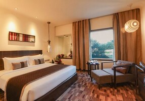 Quarto de hotel com uma cama grande, uma mesinha, uma cadeira e uma janela com cortinas.