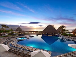 Área de piscina de resort com várias espreguiçadeiras e guarda-sóis, cercada por construções de palha, com vista para um corpo de água ao pôr do sol.