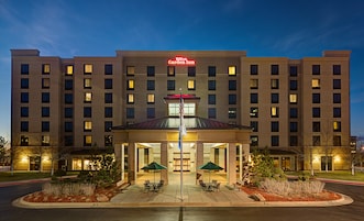 Um hotel de vários andares com uma placa grande de 'Hilton Garden Inn', uma entrada coberta com assentos e um mastro com a bandeira americana.