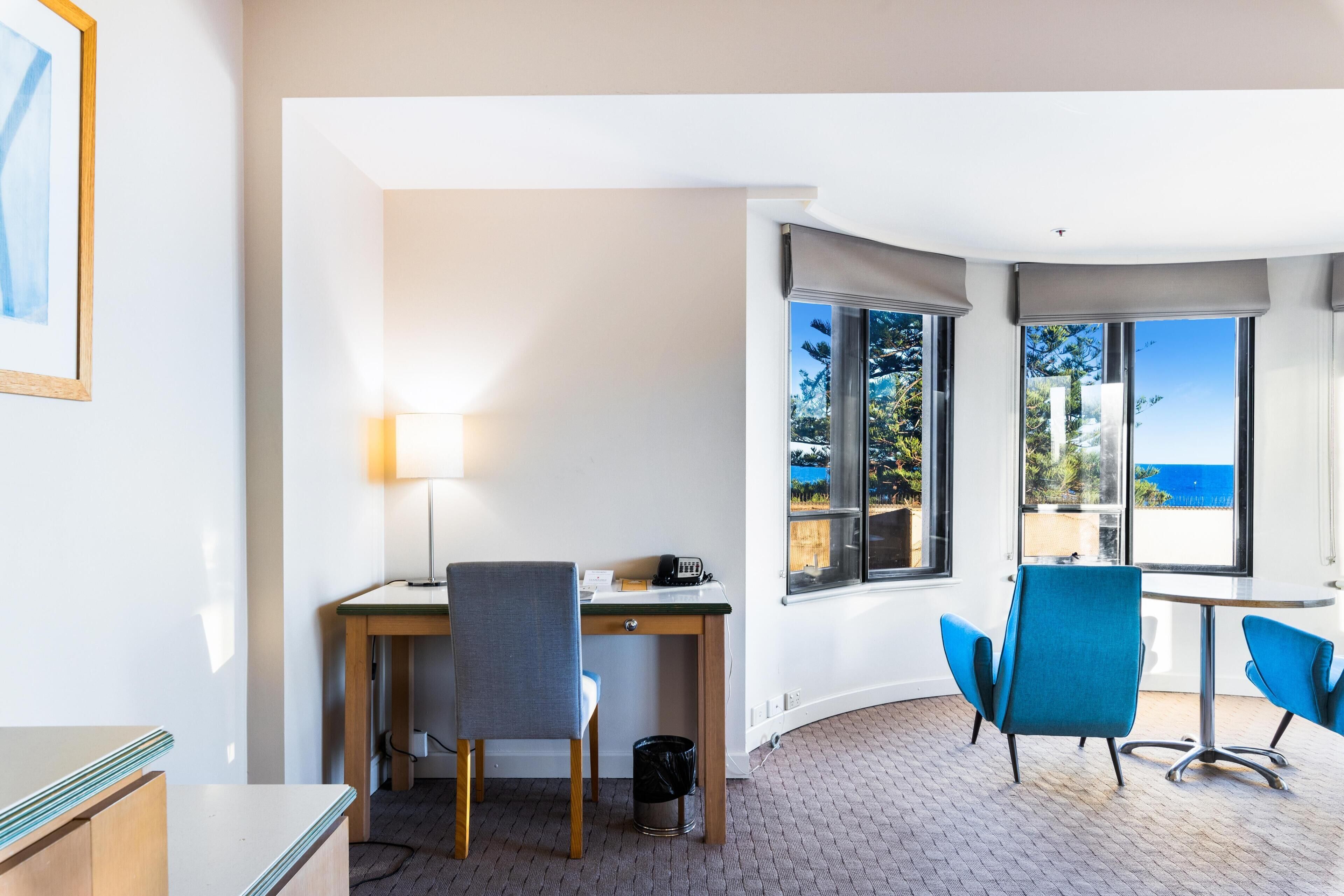 Quarto de hotel com escrivaninha, duas cadeiras, luminária, lixeira e vista para o mar.