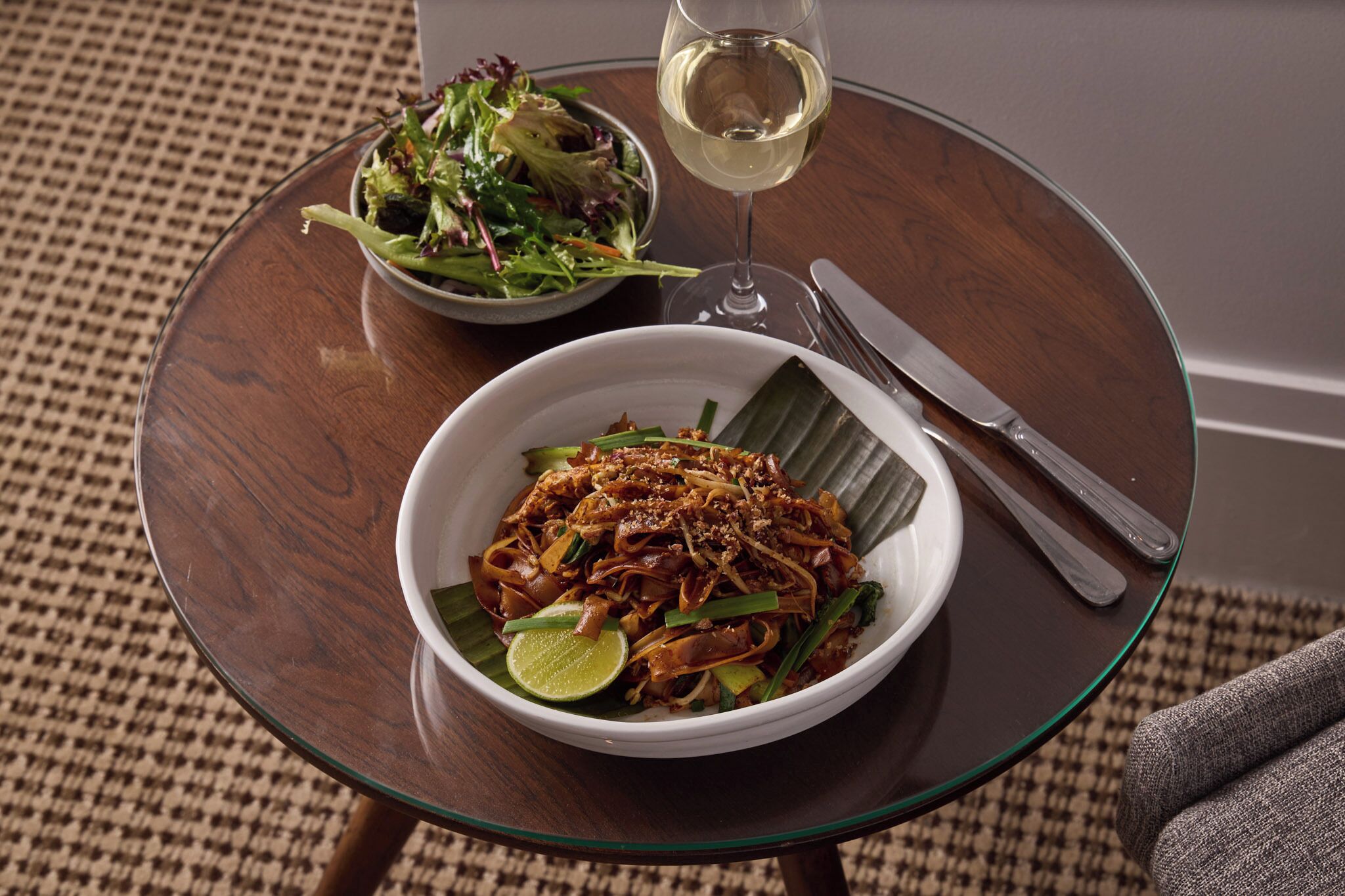 Uma mesa com uma tigela de Pad Thai, uma salada e um copo de vinho branco.