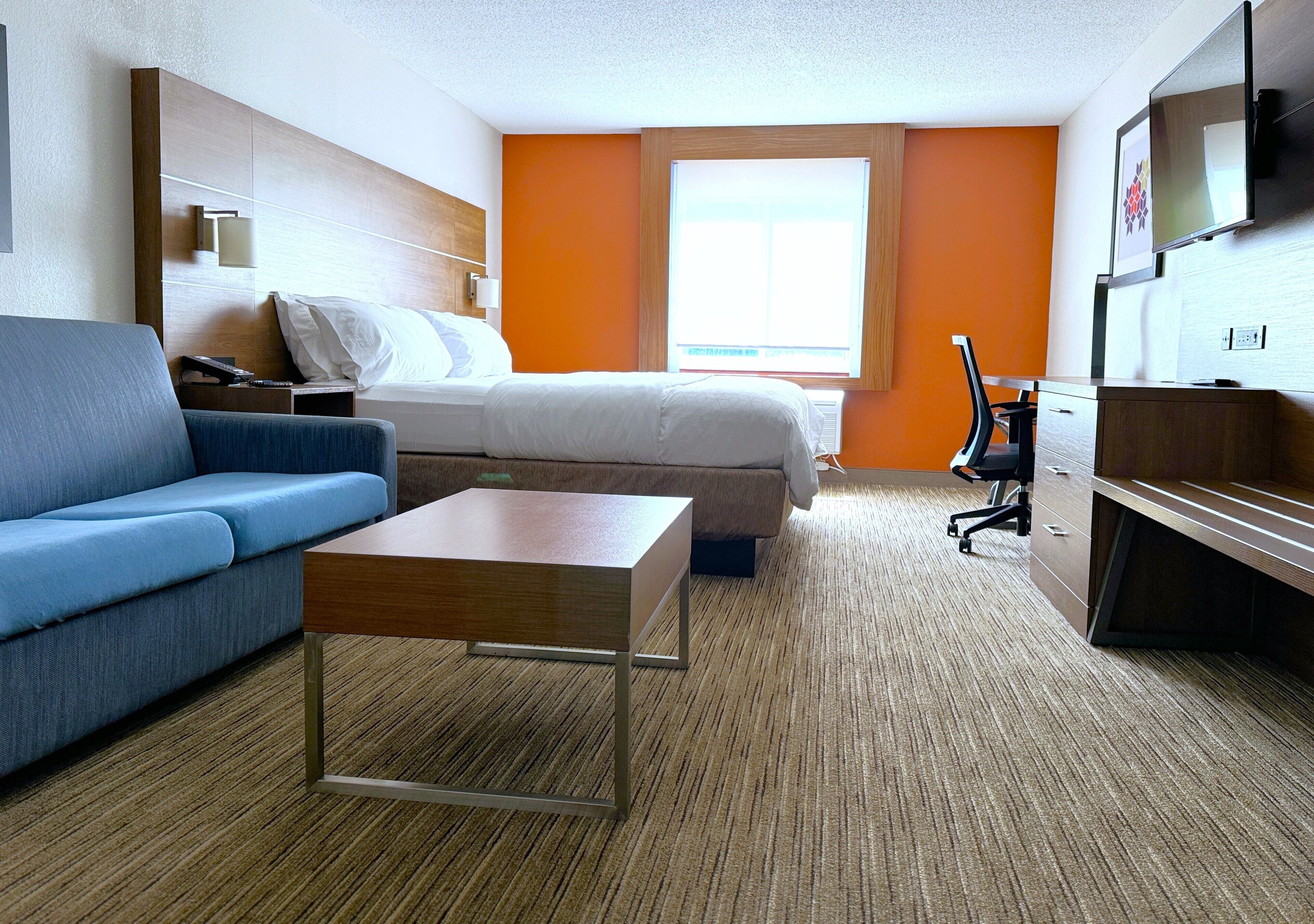 Quarto de hotel com cama, sofá azul, mesa de centro de madeira, escrivaninha e televisão.