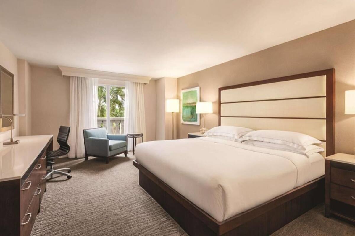 Quarto de hotel com uma cama grande, uma escrivaninha com cadeira, uma mesinha e uma janela com cortinas.