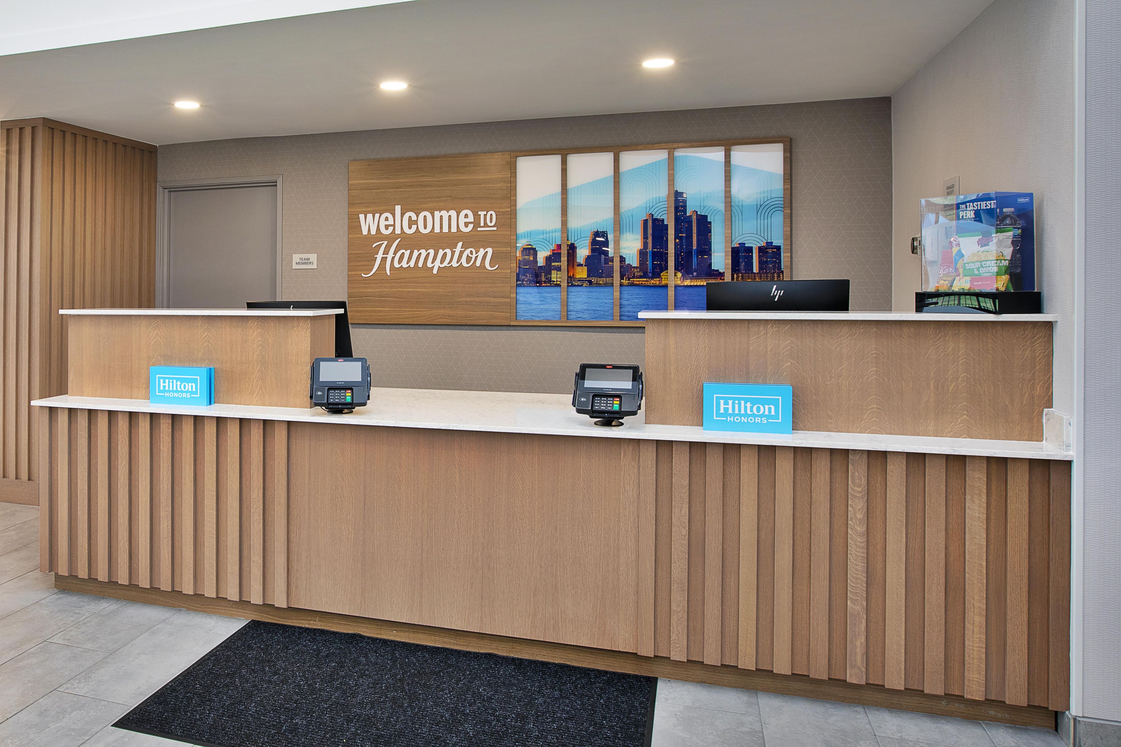 Balcão de recepção de hotel com um letreiro 'Welcome to Hampton', um cartaz com vista da cidade e um expositor de cartão de fidelidade 'Hilton Honors'.