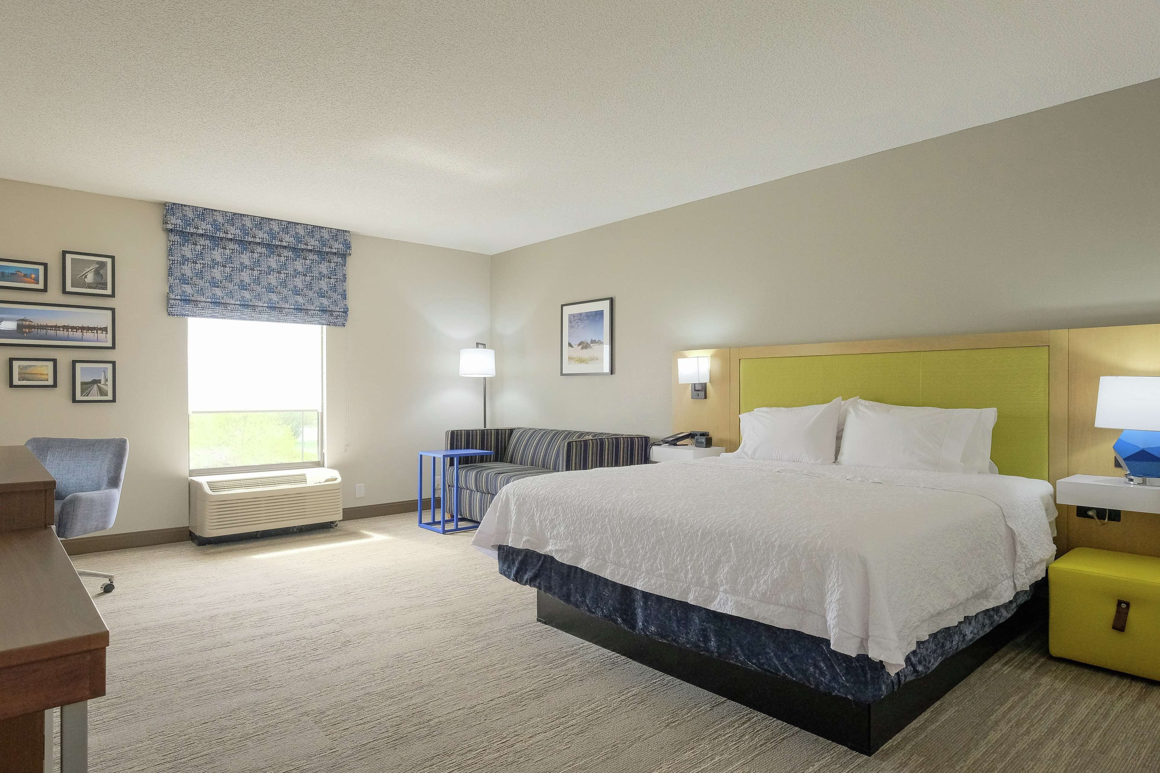 Quarto de hotel com uma cama grande, uma escrivaninha, uma cadeira, uma janela com persianas e um ar condicionado fixado na parede.