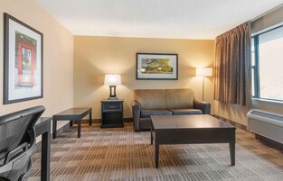Quarto de hotel com sofá, mesa pequena, cadeira, luminária, quadro na parede e janela com cortinas.
