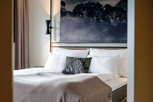 Cama bem arrumada com almofadas brancas e estampadas, cabeceira escura e uma pintura de paisagem emoldurada acima.