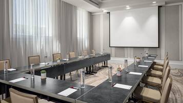 Uma sala de conferências com uma mesa comprida, cadeiras, tela de projetor e uma grande janela com cortinas.