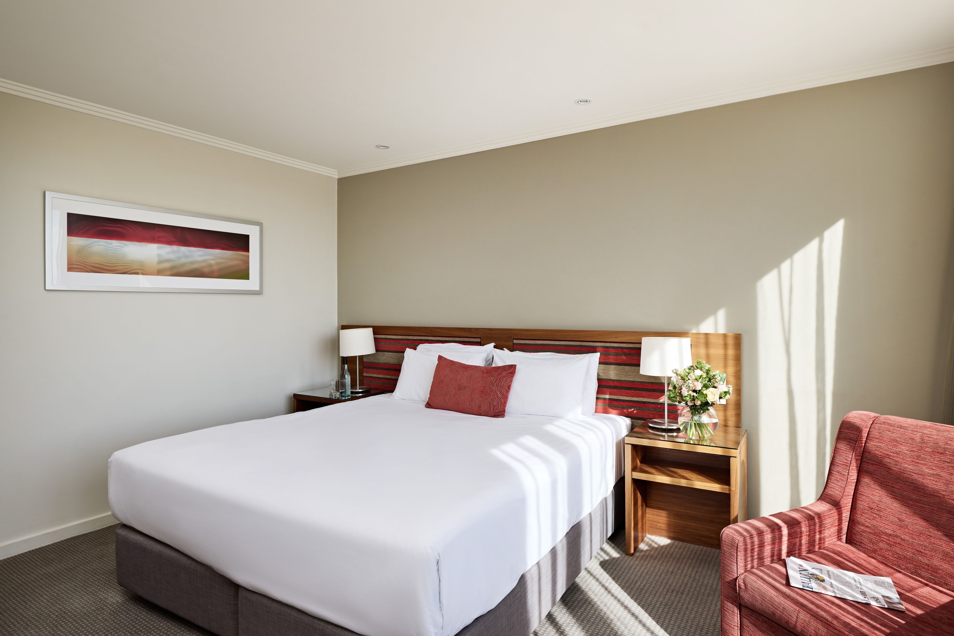 Quarto de hotel com uma cama grande, uma mesa de cabeceira de madeira, uma cadeira vermelha e um quadro na parede.