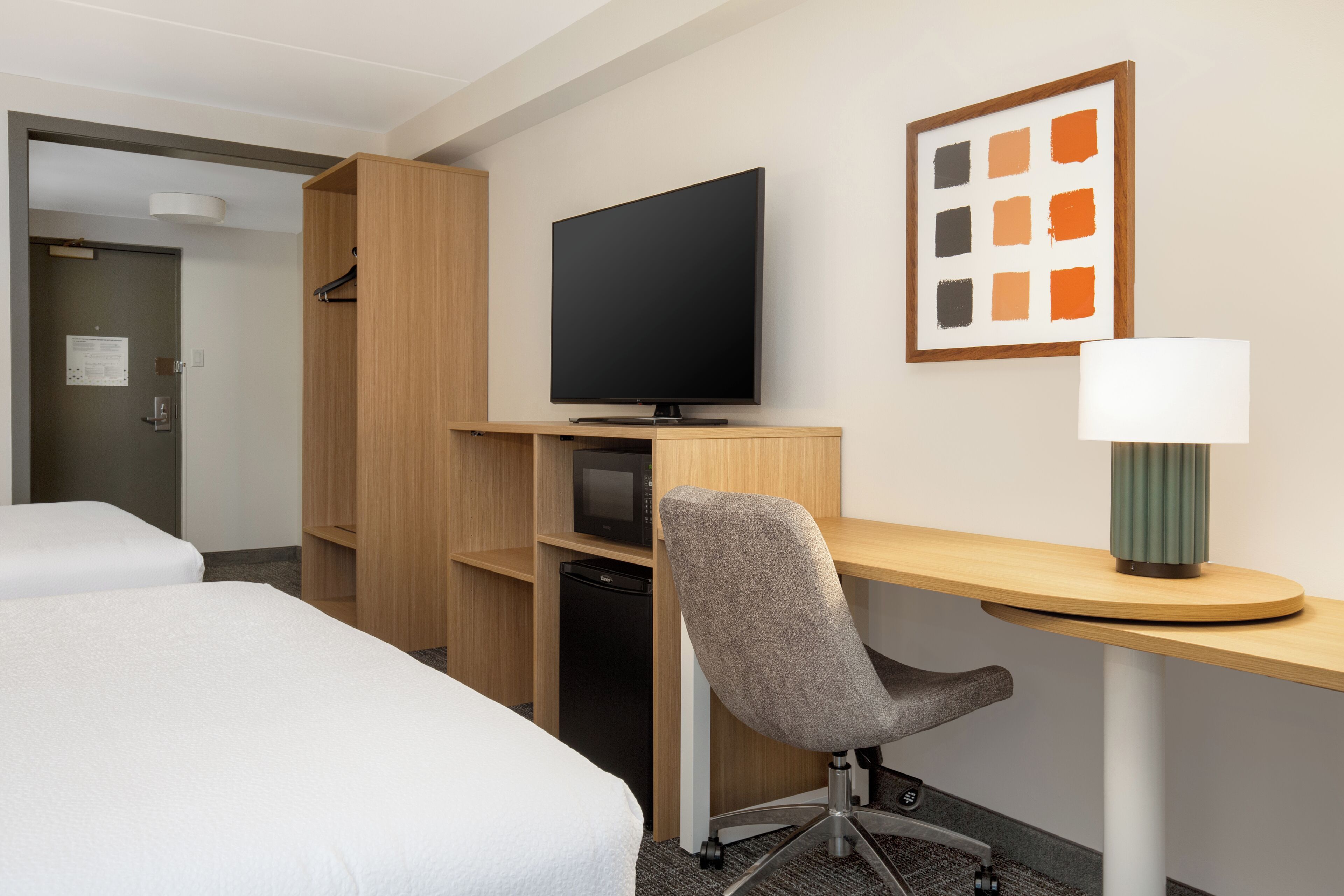 Quarto de hotel com uma cama, escrivaninha com cadeira, televisão, micro-ondas e frigorífico.
