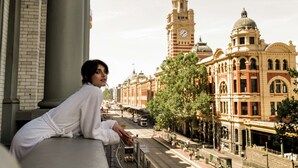 Uma pessoa apoiada em uma varanda com vista para um horizonte urbano com edifícios históricos e um campanário.