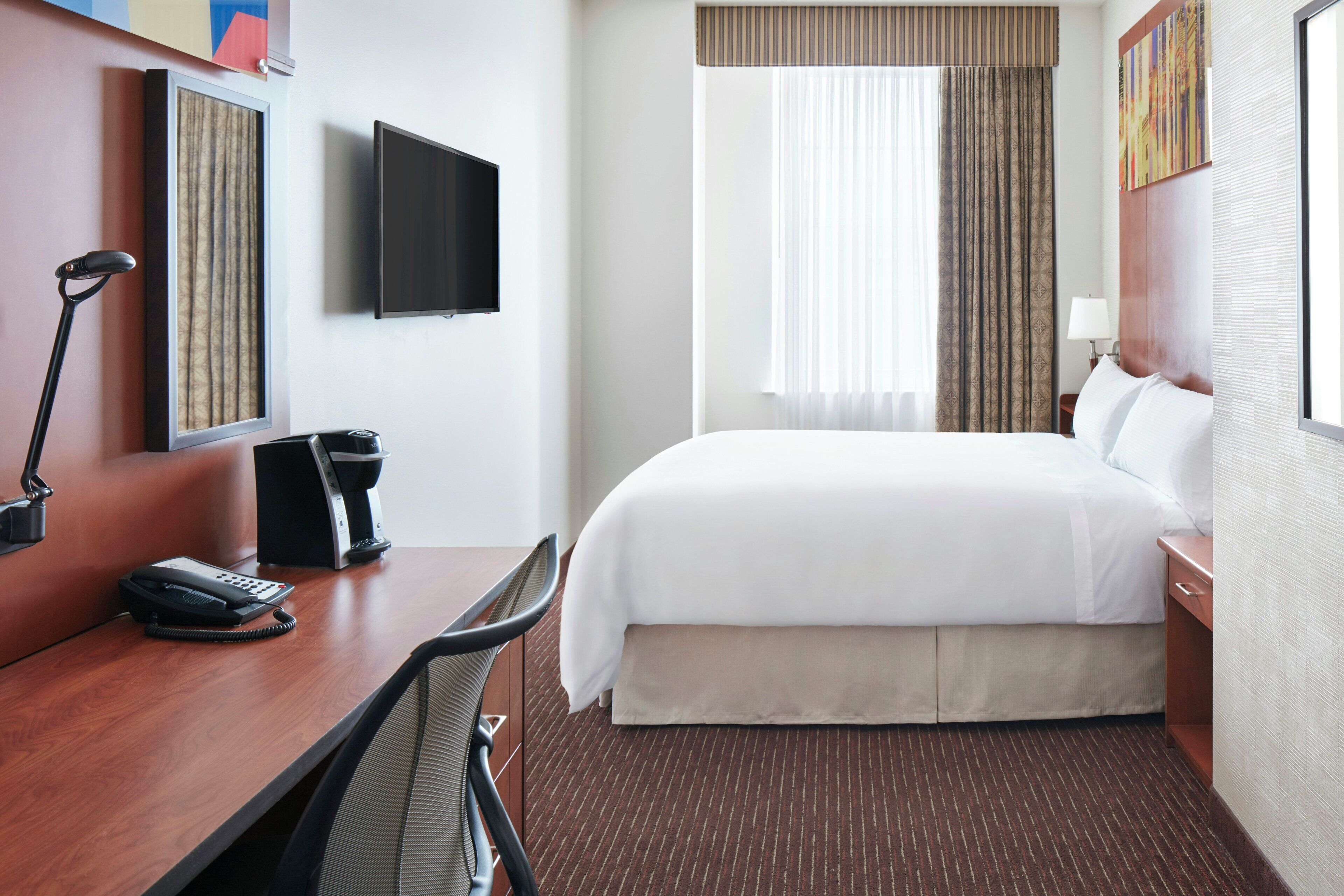 Quarto de hotel com cama, escrivaninha, cadeira, telefone, televisão e cortinas.