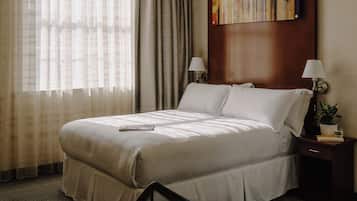 Quarto de hotel com uma cama grande, duas luminárias de cabeceira, cabeceira com obra de arte, escrivaninha com cadeira e janela com cortinas translúcidas.