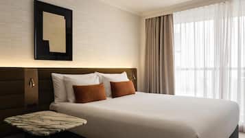 Quarto de hotel com uma cama grande, uma mesinha, um banquinho redondo, um quadro na parede e uma luminária no teto.