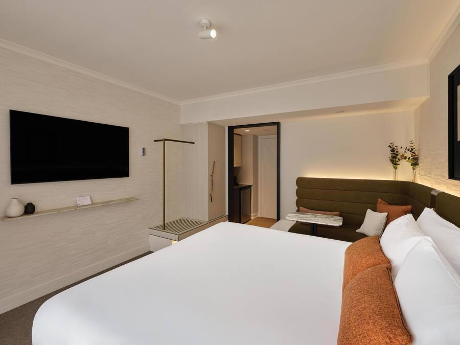 Quarto de hotel com uma cama grande, televisão de tela plana na parede, mesa de cabeceira e porta de acesso a um banheiro.