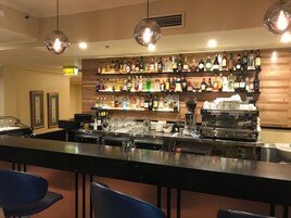 Uma área de bar com um balcão comprido, várias prateleiras com garrafas de bebidas e uma máquina de café.