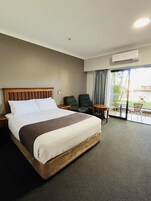 Quarto de hotel com uma cama grande, uma mesinha, duas cadeiras, aparelho de ar condicionado e uma porta de vidro deslizante para uma área externa.