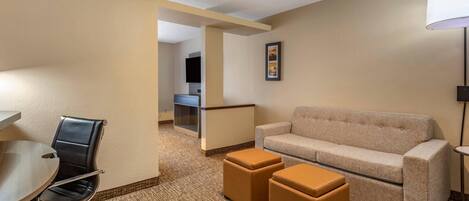 Quarto de hotel com sofá, dois pufes, escrivaninha com cadeira, TV de tela plana na parede e luminária.