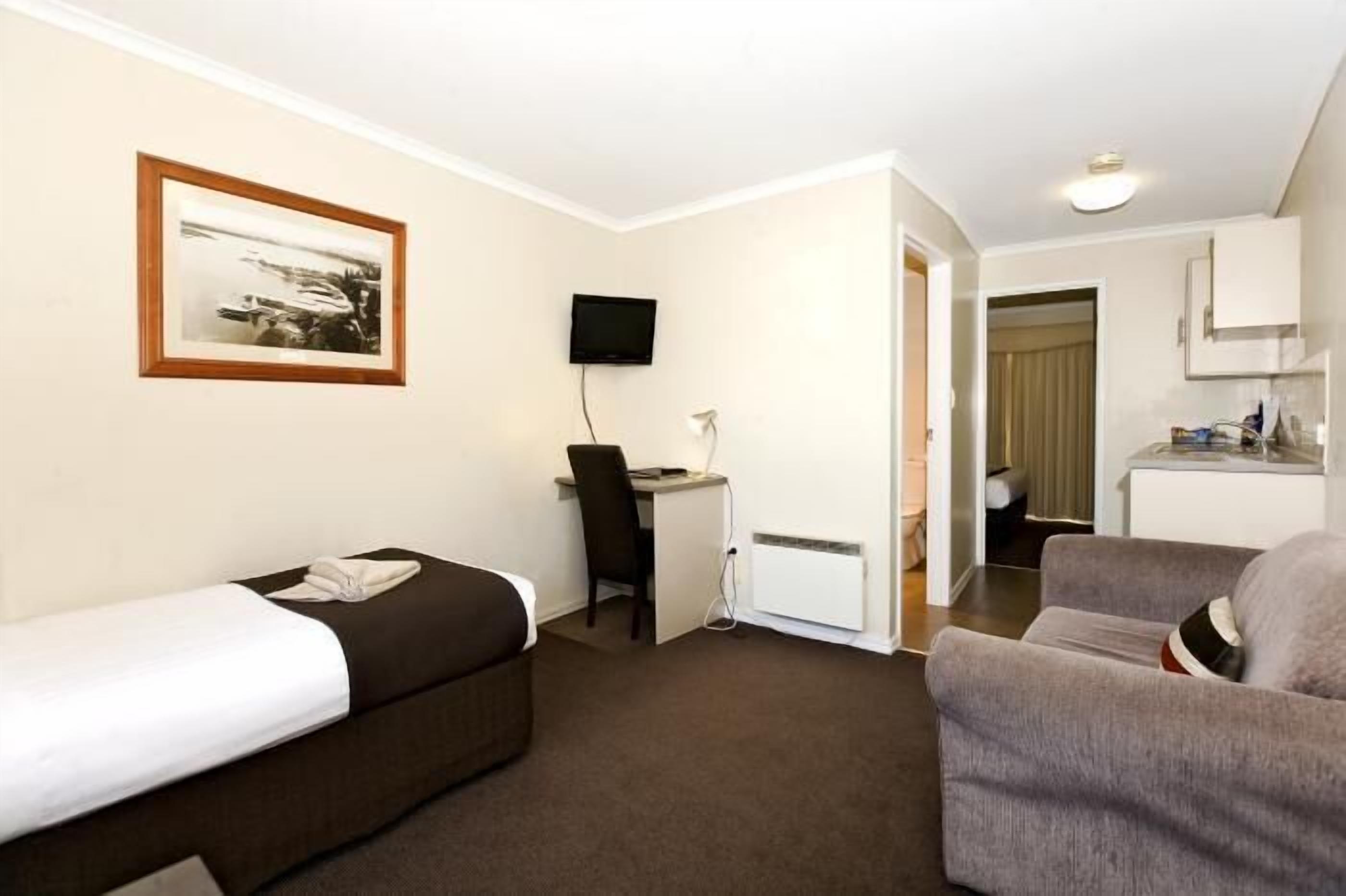 Quarto de hotel com uma cama, uma escrivaninha, uma cadeira, televisão e um quadro na parede.