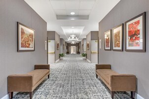 Um corredor de hotel com carpete estampado, quadros nas paredes e duas áreas de estar com bancos estofados.