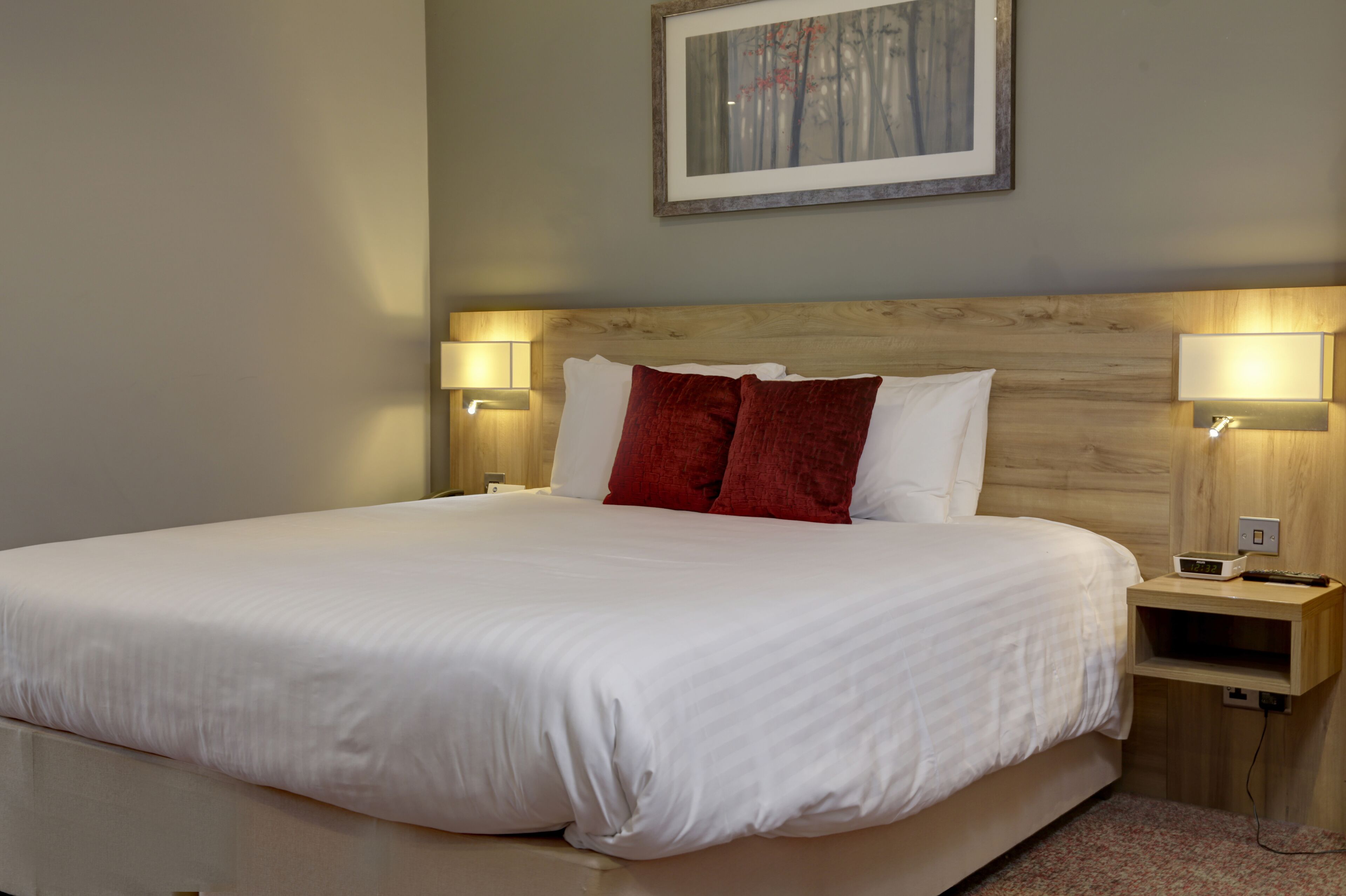 Cama bem arrumada com travesseiros brancos e vermelhos, cabeceira de madeira e abajures laterais.