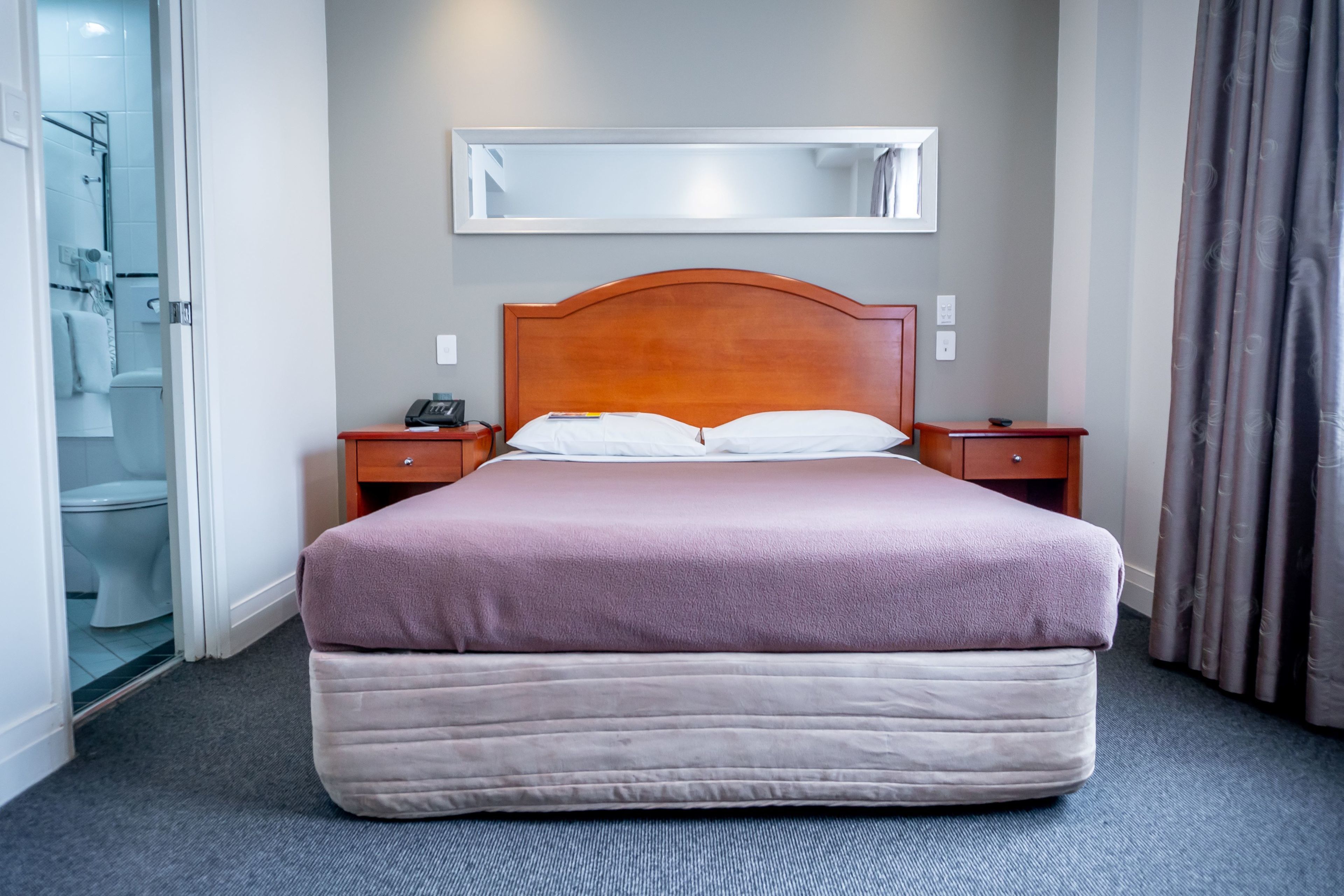 Quarto de hotel com cama grande, mesa de cabeceira, espelho acima da cama e banheiro visível por uma porta aberta.