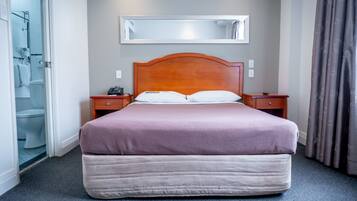 Quarto de hotel com cama grande, mesa de cabeceira, espelho acima da cama e banheiro visível por uma porta aberta.