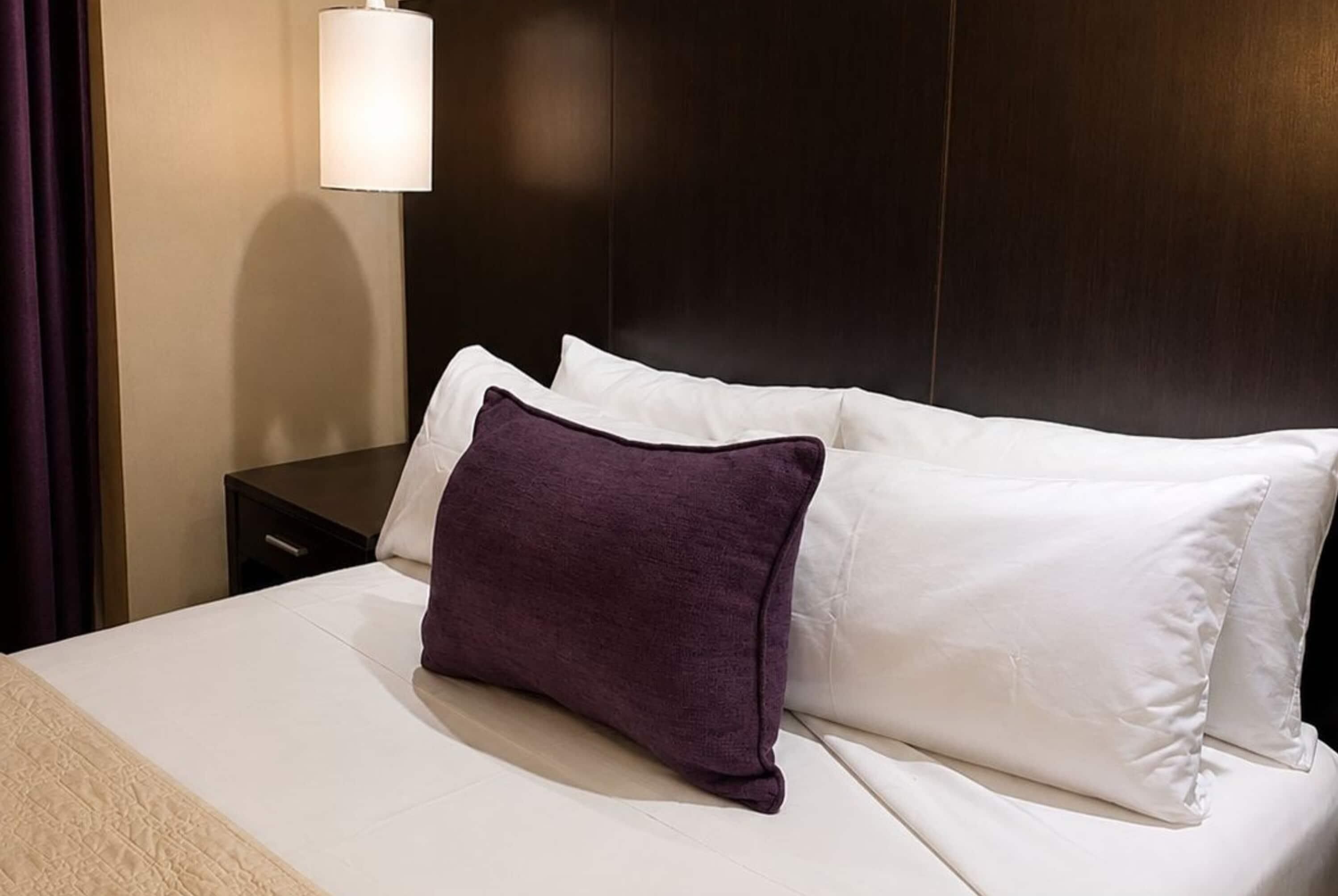 Quarto de hotel com uma cama, mesa de cabeceira, abajur e uma parede escura.
