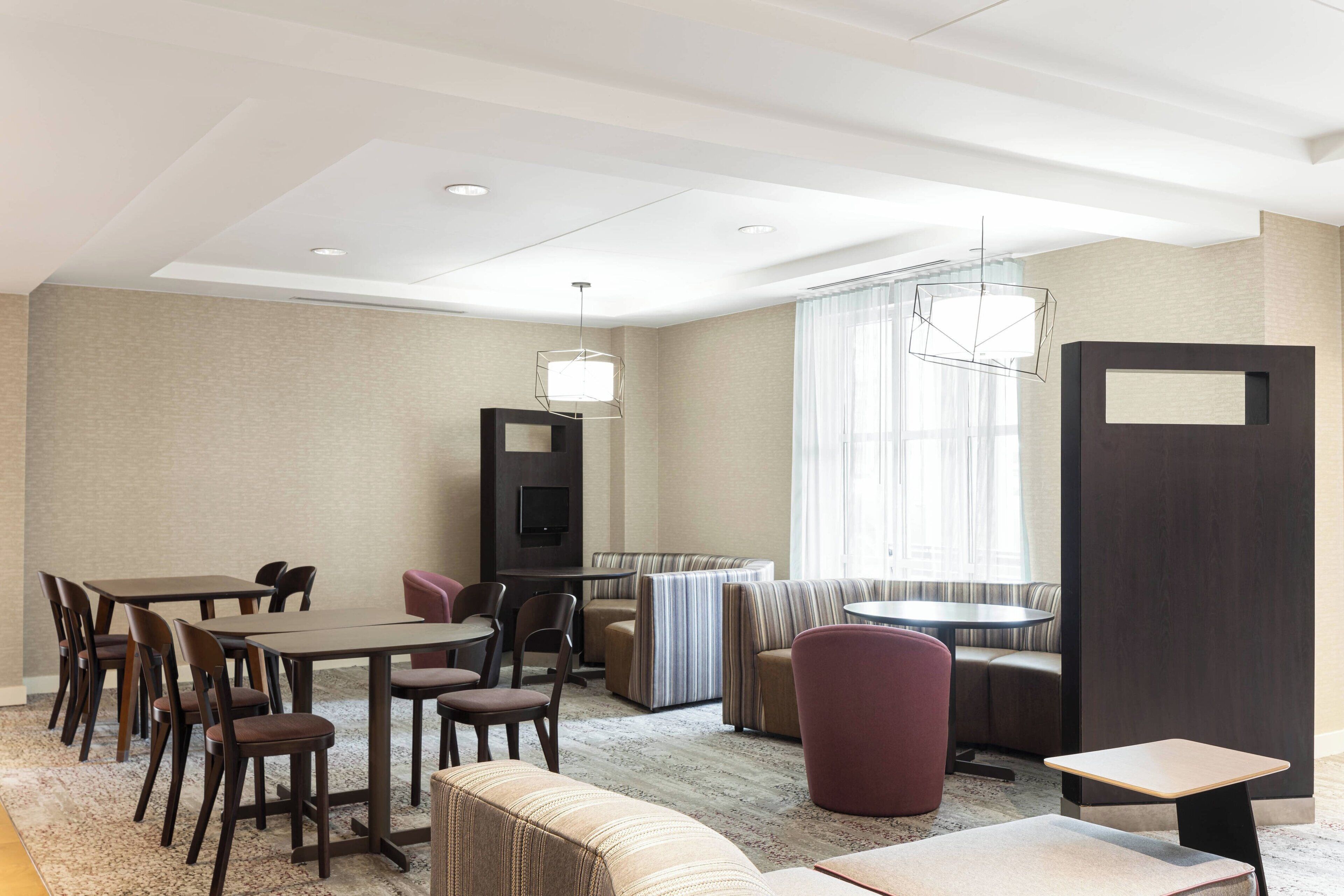 Um saguão de hotel com área de refeições, assentos e uma grande janela com cortinas.
