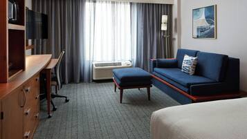 Quarto de hotel com escrivaninha, sofá azul, cama, televisão, ar-condicionado e janela com cortinas.