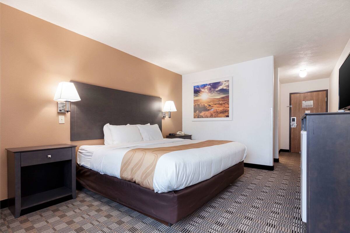 Quarto de hotel com uma cama grande, duas mesas de cabeceira, um criado-mudo, uma luminária na parede, um quadro e uma porta que dá para outro cômodo.