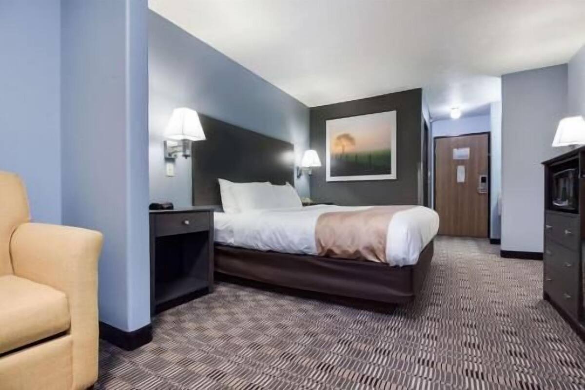 Quarto de hotel com cama, abajures, escrivaninha, cadeira, guarda-roupa e um quadro na parede.