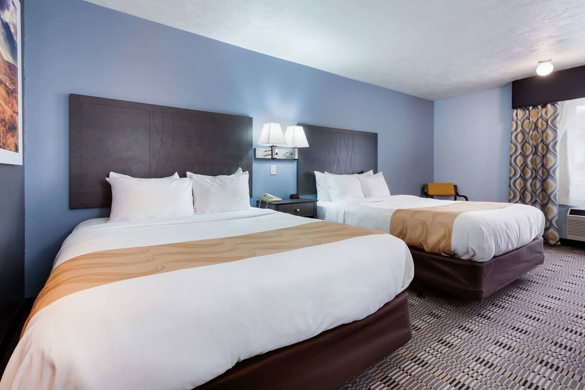 Quarto de hotel com duas camas, uma mesa de cabeceira, um abajur e uma janela com cortinas.