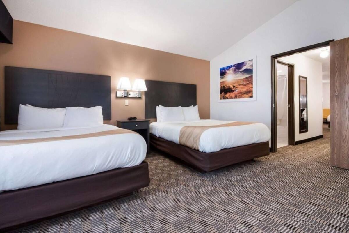 Quarto de hotel com duas camas, uma mesa de cabeceira com abajures, um quadro na parede e uma porta que dá para outro aposento.