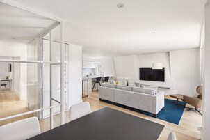 Uma sala de estar moderna com um sofá cinzento, uma mesa de jantar e uma cozinha ao fundo.