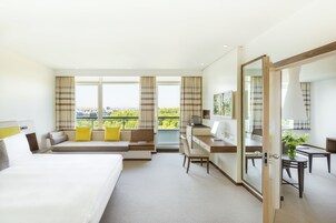 Quarto de hotel com uma cama grande, um sofá com almofadas amarelas, uma escrivaninha com cadeira e vista da cidade por grandes janelas.