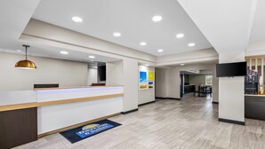 Uma área de recepção de hotel com balcão, tela digital, iluminação embutida e vista para a sala de jantar.