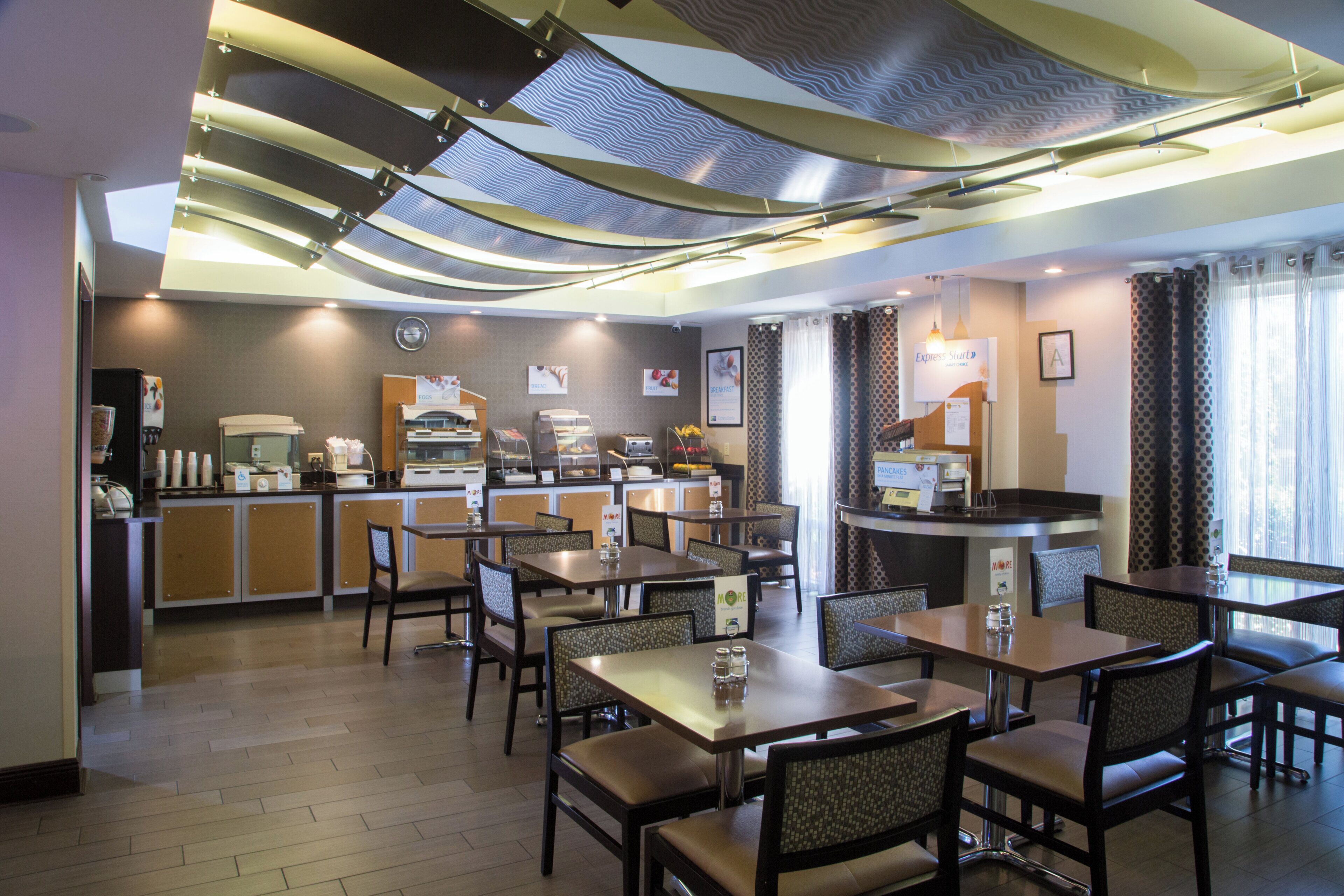 Uma sala de jantar de hotel com várias mesas e cadeiras, uma área de cozinha e um balcão de café da manhã.