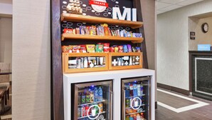 Máquina de vendas com lanches e bebidas, um letreiro com 'TREAT' e 'MI', e um relógio na parede.