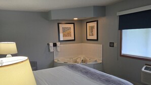 Quarto de hotel com cama, mesa de cabeceira com abajur, banheiro com banheira e quadros nas paredes.