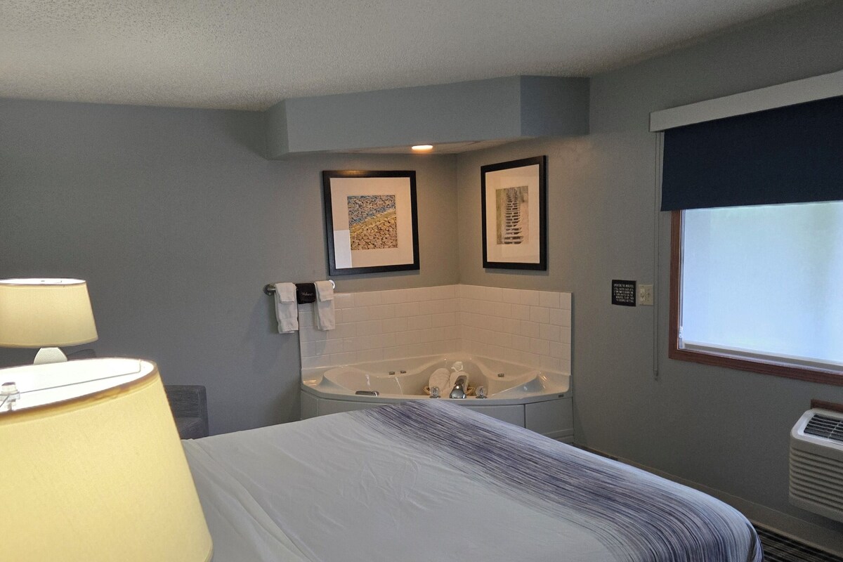 Quarto de hotel com cama, mesa de cabeceira com abajur, banheiro com banheira e quadros nas paredes.