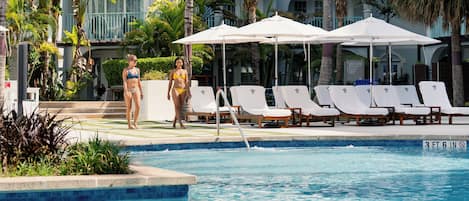 Área da piscina com espreguiçadeiras e guarda-sóis, um letreiro indicando as comodidades, e um edifício com varandas ao fundo.