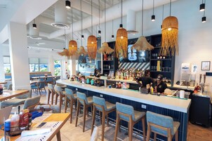 Uma área de bar com banquetas altas, um balcão com vários tipos de bebidas e luminárias de vime suspensas.