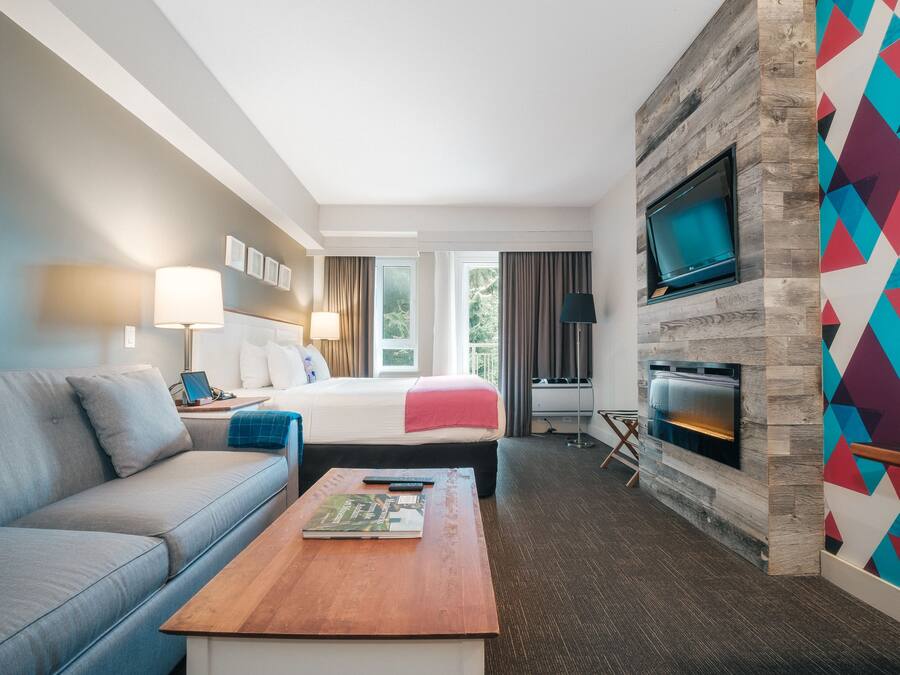 Quarto de hotel com cama, sofá, mesa de centro, televisão, lareira e janela com cortinas.