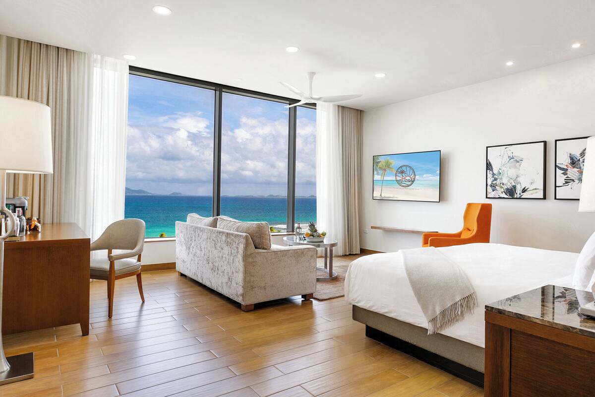 Quarto de hotel com uma grande janela que oferece vista para o mar, uma cama, uma escrivaninha, uma cadeira e uma pequena mesa com uma tigela de frutas.