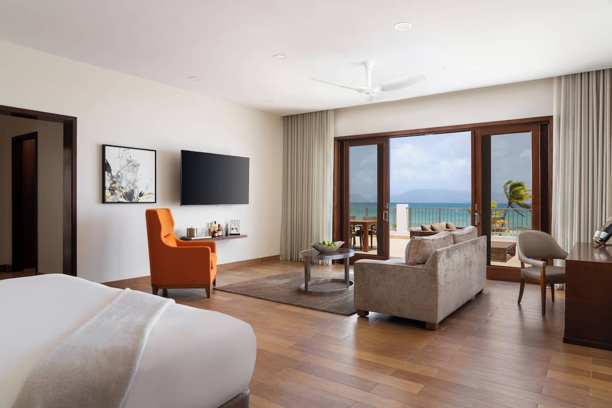 Quarto de hotel espaçoso com cama grande, sofá, mesa de jantar e TV na parede. O quarto possui varanda vista para o oceano.