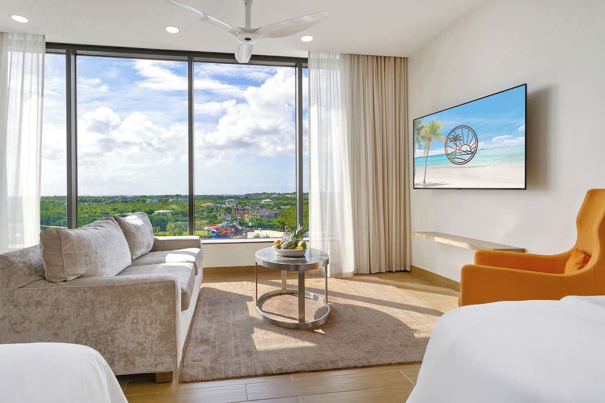 Quarto de hotel moderno com uma janela grande, um sofá, uma cama, uma cadeira e uma pequena mesa com frutas.